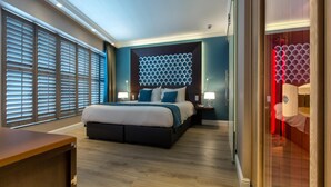 Quarto de hotel moderno com uma cama grande, duas mesas de cabeceira, luminária fixada na parede e vista para o exterior através de amplas janelas com persianas.