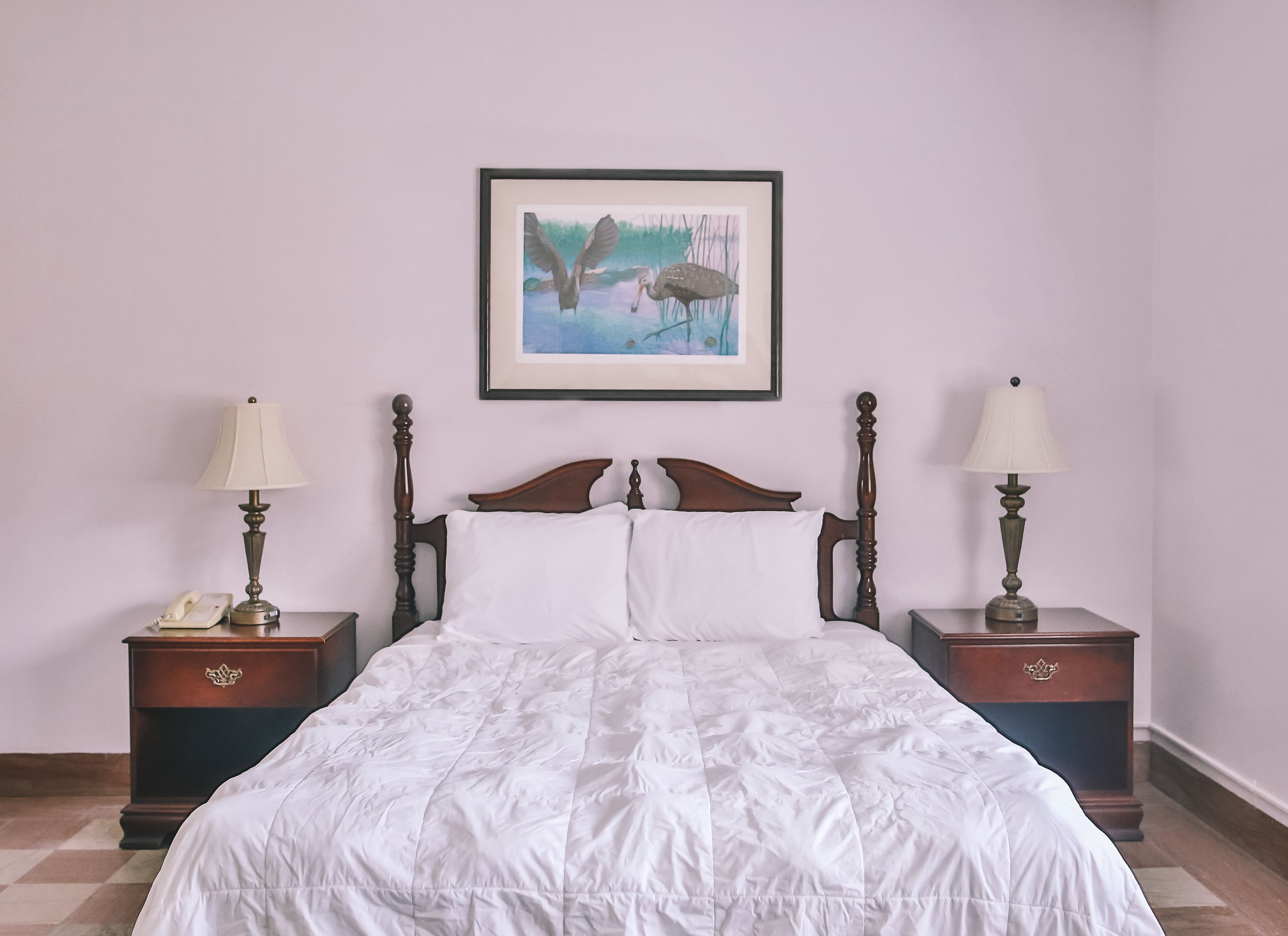 Uma cama bem arrumada com roupa de cama branca, com duas mesas de cabeceira com luminárias, um quadro emoldurado acima da cama e um chão xadrez.