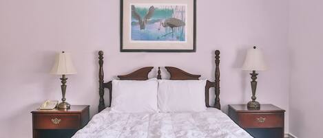 Uma cama bem arrumada com roupa de cama branca, com duas mesas de cabeceira com luminárias, um quadro emoldurado acima da cama e um chão xadrez.