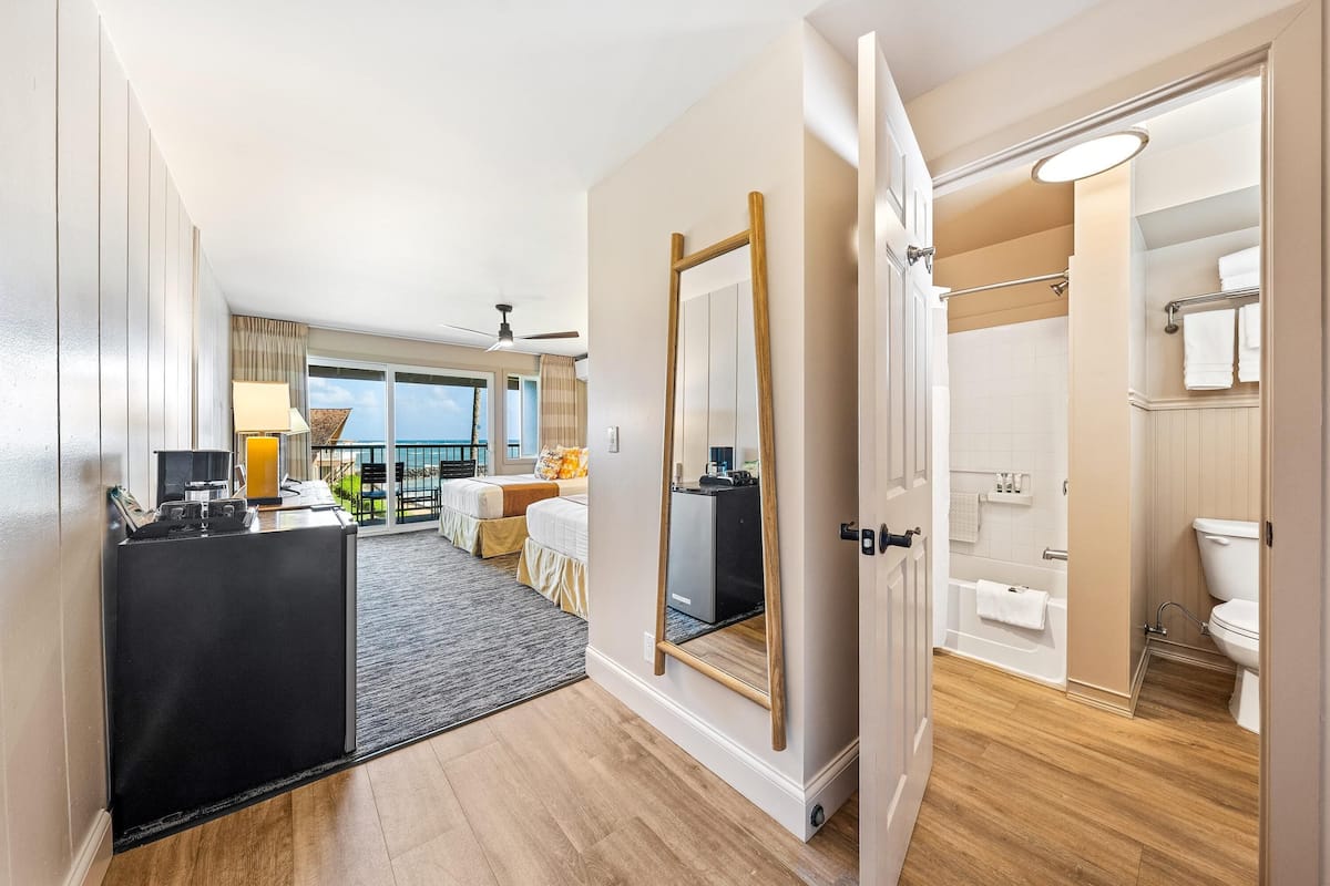 Quarto de hotel moderno com cozinha compacta, um espelho grande, banheiro com banheira e vaso sanitário, e vista para o oceano pela varanda.