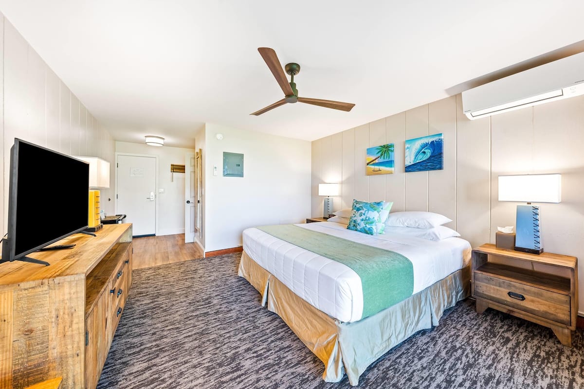 Quarto de hotel com cama, cômoda de madeira, televisão, ventilador de teto e obras de arte nas paredes.