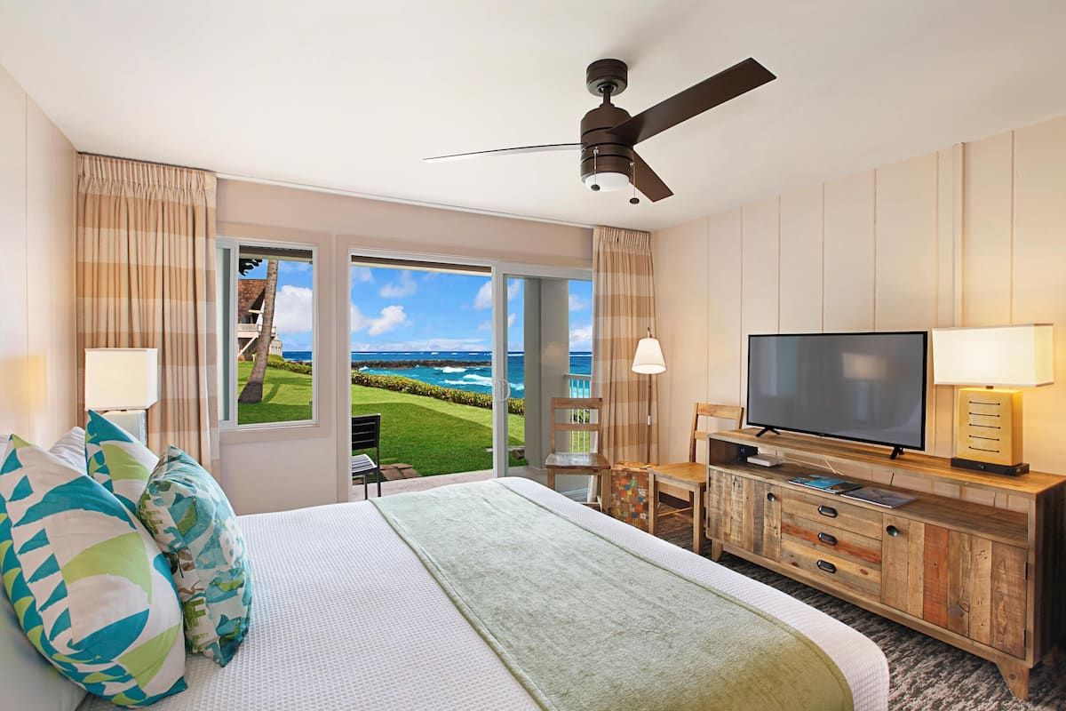 Quarto de hotel com uma cama grande, televisão, cômoda de madeira e varanda com vista para o mar.