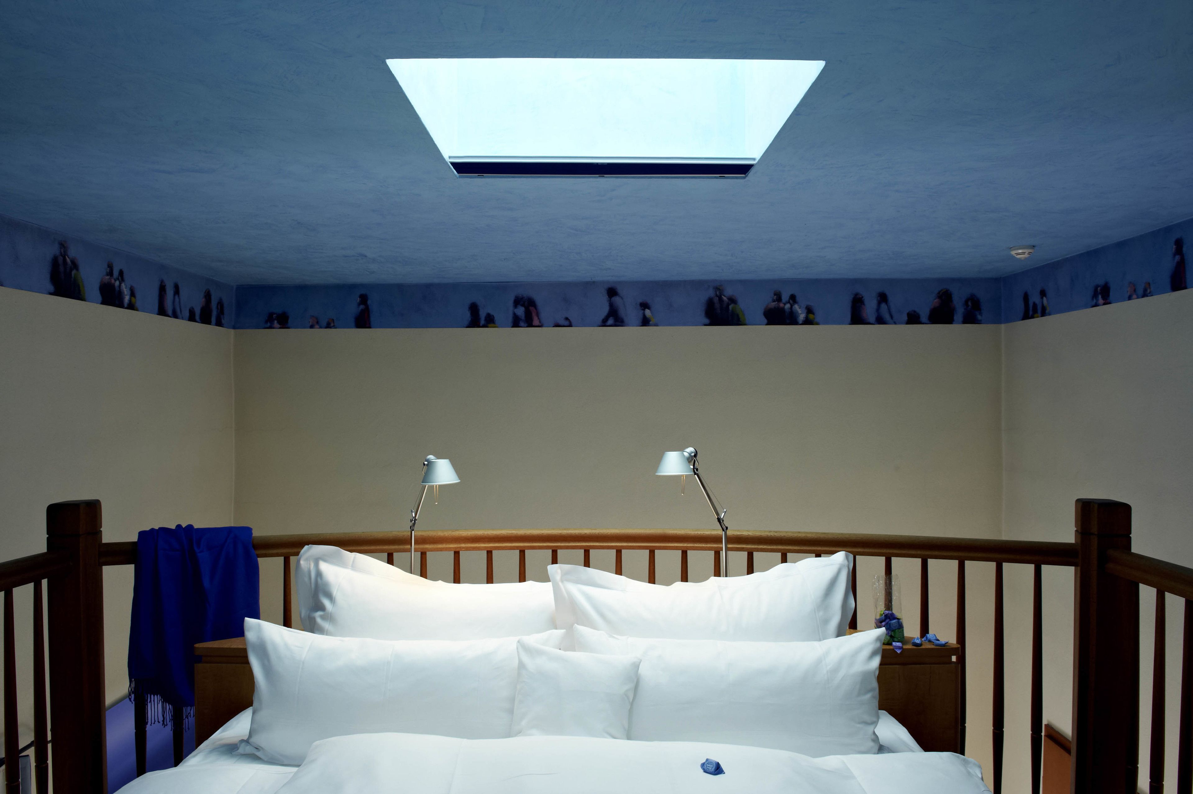 Quarto com claraboia, cama com roupa de cama branca, duas luminárias de cabeceira e uma toalha azul no lado esquerdo.