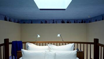 Quarto com claraboia, cama com roupa de cama branca, duas luminárias de cabeceira e uma toalha azul no lado esquerdo.