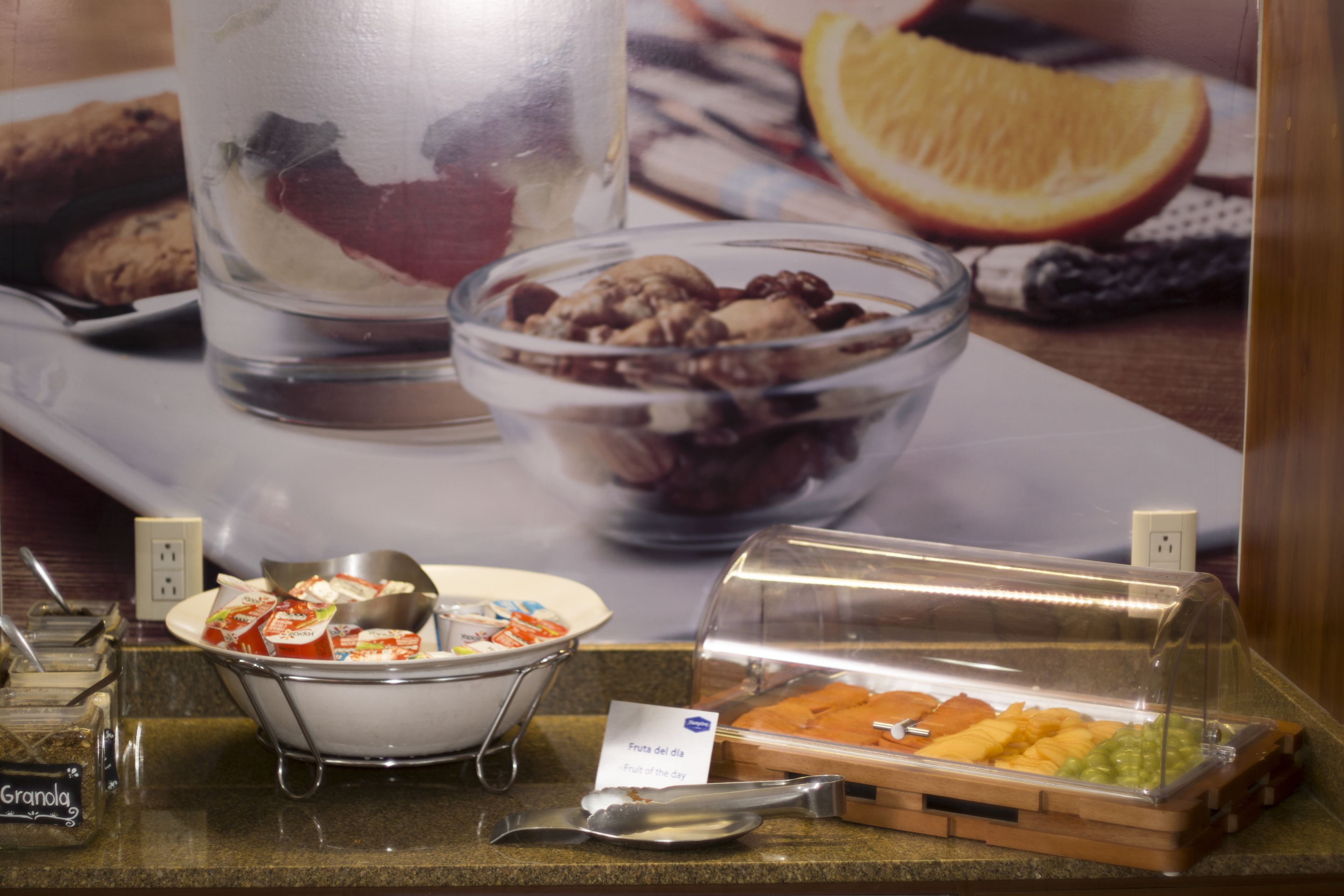 Buffet com diversos alimentos, incluindo uma tigela de granola, um copo de chá gelado e uma vitrine com frutas e doces.