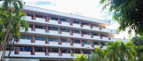 Um hotel de vários andares com entrada central, rodeado por palmeiras e um estacionamento.