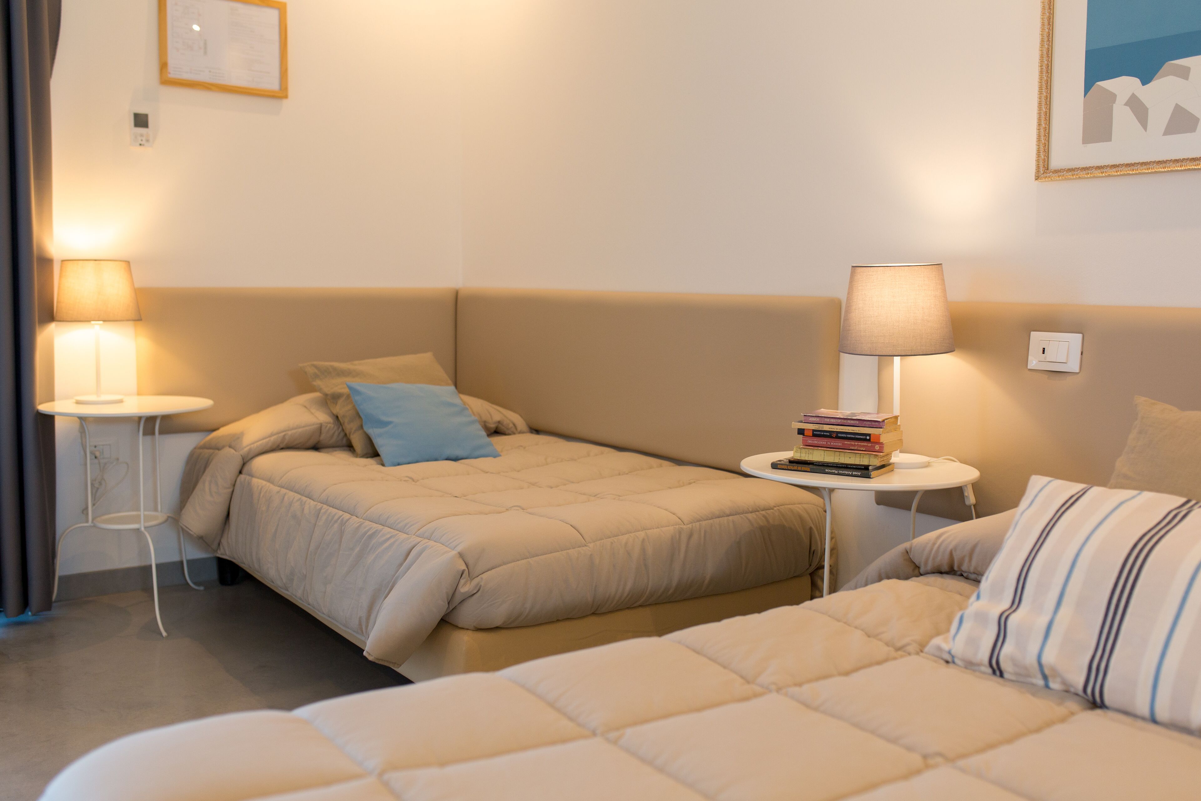 Quarto de hotel com duas camas, cada uma com travesseiro e mesa de cabeceira com abajur. Há um quadro na parede e uma pequena mesa com livros.