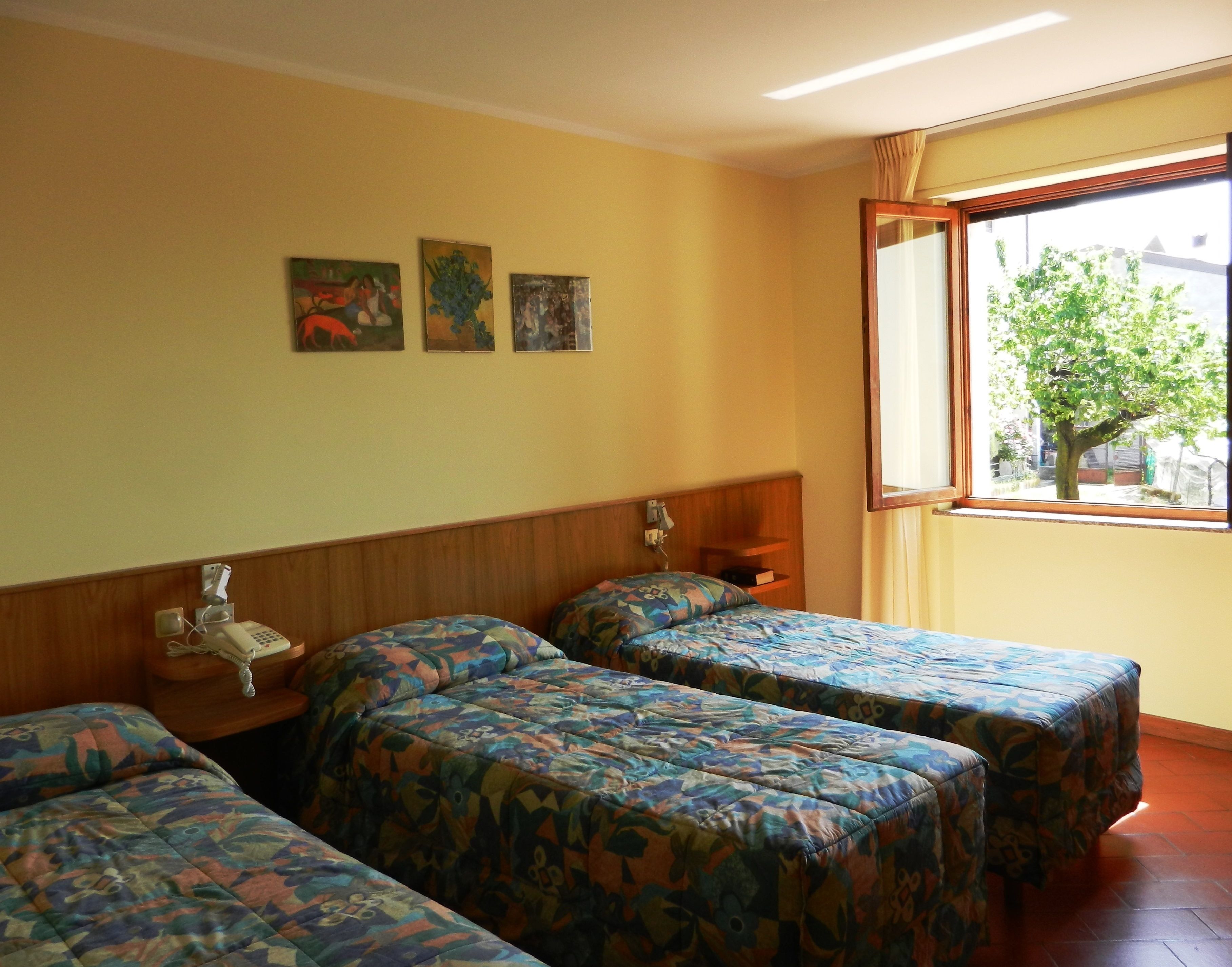 Quarto de hotel com duas camas de solteiro, cabeceira de madeira, mesa de cabeceira com abajur e janela com vista para uma árvore e edifícios.