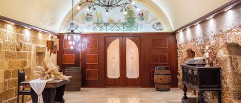 Quarto com parede de pedra, porta de madeira com painéis decorativos, um candelabro e uma mesa com toalha branca.