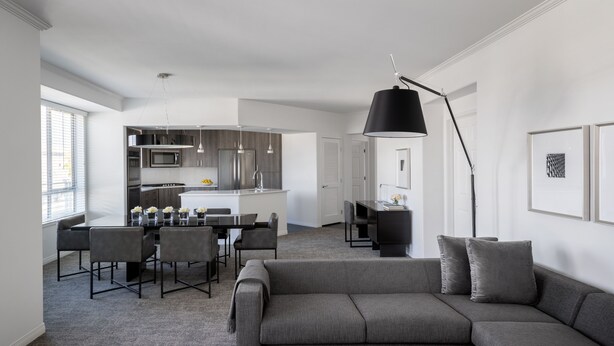 Uma sala moderna com um sofá seccional cinza, uma área de jantar com mesa e cadeiras, e uma cozinha com eletrodomésticos de aço inoxidável.