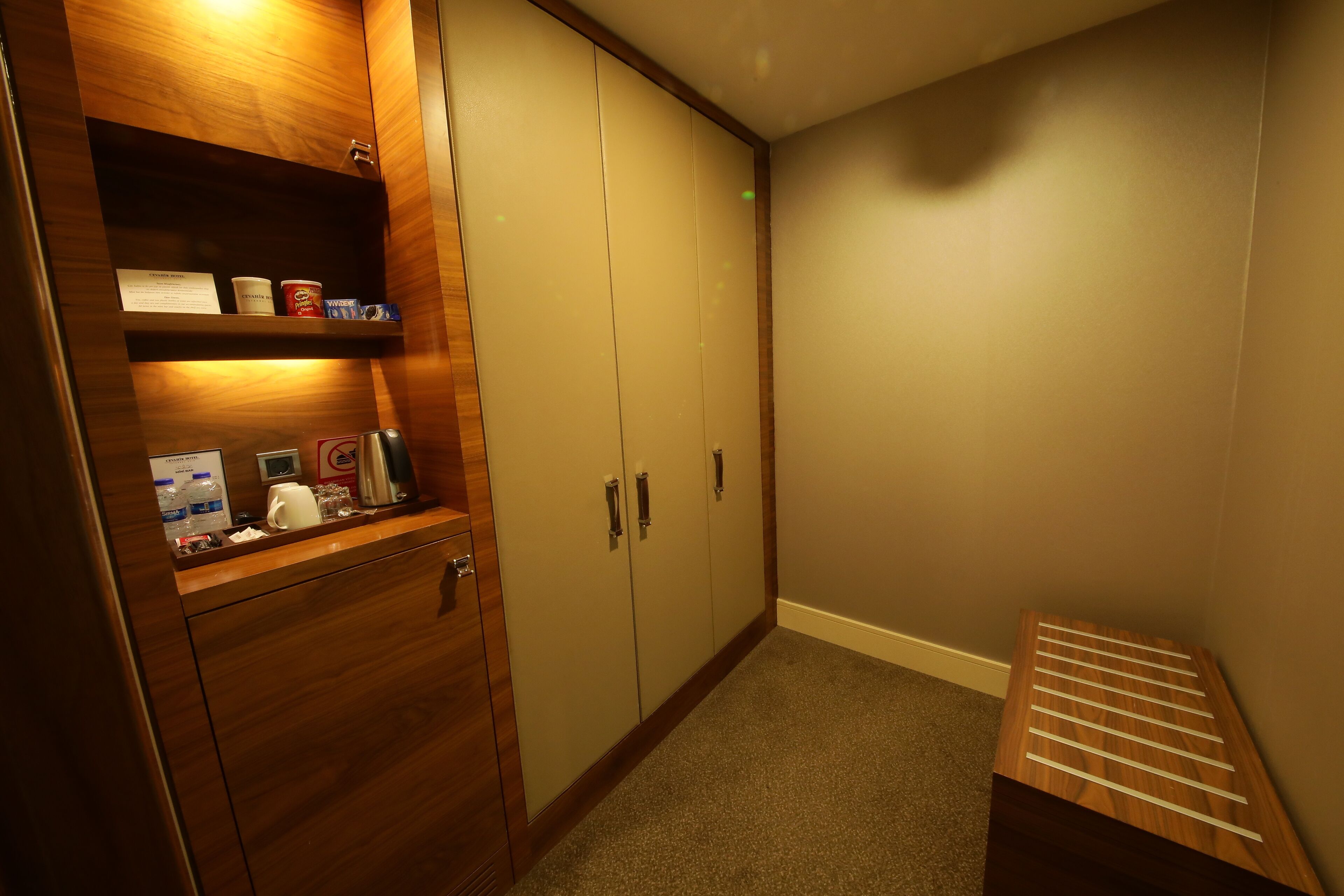 Quarto de hotel com guarda-roupa de madeira, um pequeno espaço com produtos de higiene e um banco de madeira.