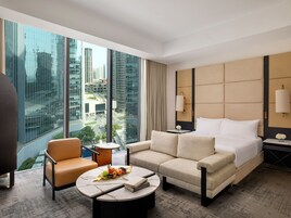 Quarto de hotel moderno com uma cama grande, um sofá, uma mesa de centro e vista para a cidade através de janelas do chão ao teto.