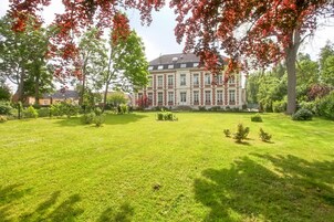 Um edifício grande e histórico com fachada simétrica, rodeado por um jardim bem cuidado e árvores maduras.