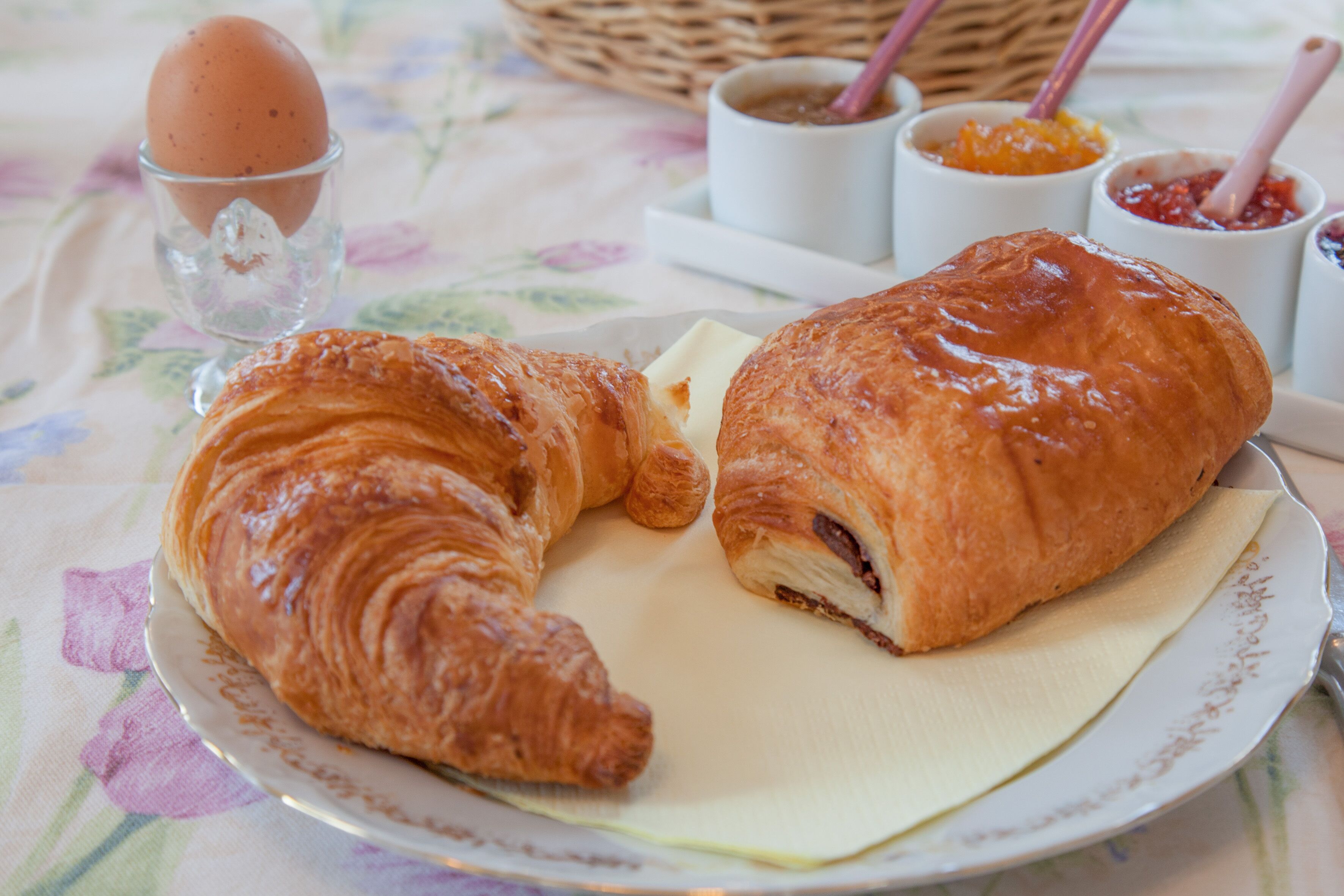 Um prato com dois croissants e uma fatia de queijo, acompanhado por um pequeno copo com ovos e três pequenos potes de geleia.