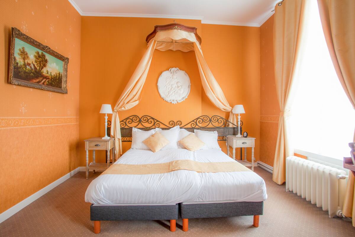 Uma cama grande com dossel, duas mesas de cabeceira, um relógio de parede e um quadro na parede.
