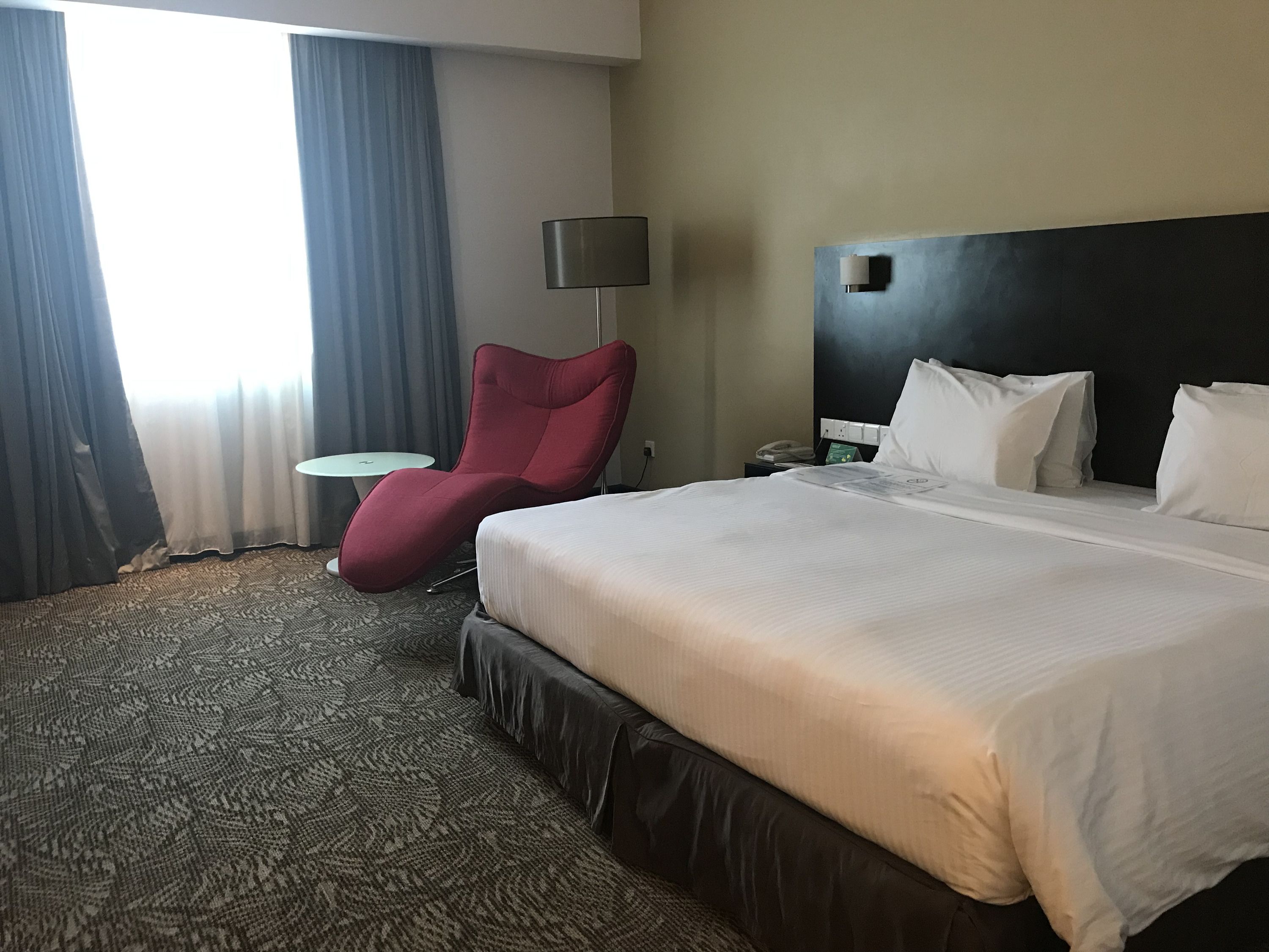 Quarto de hotel com uma cama grande, uma poltrona vermelha, uma mesa pequena e um abajur.