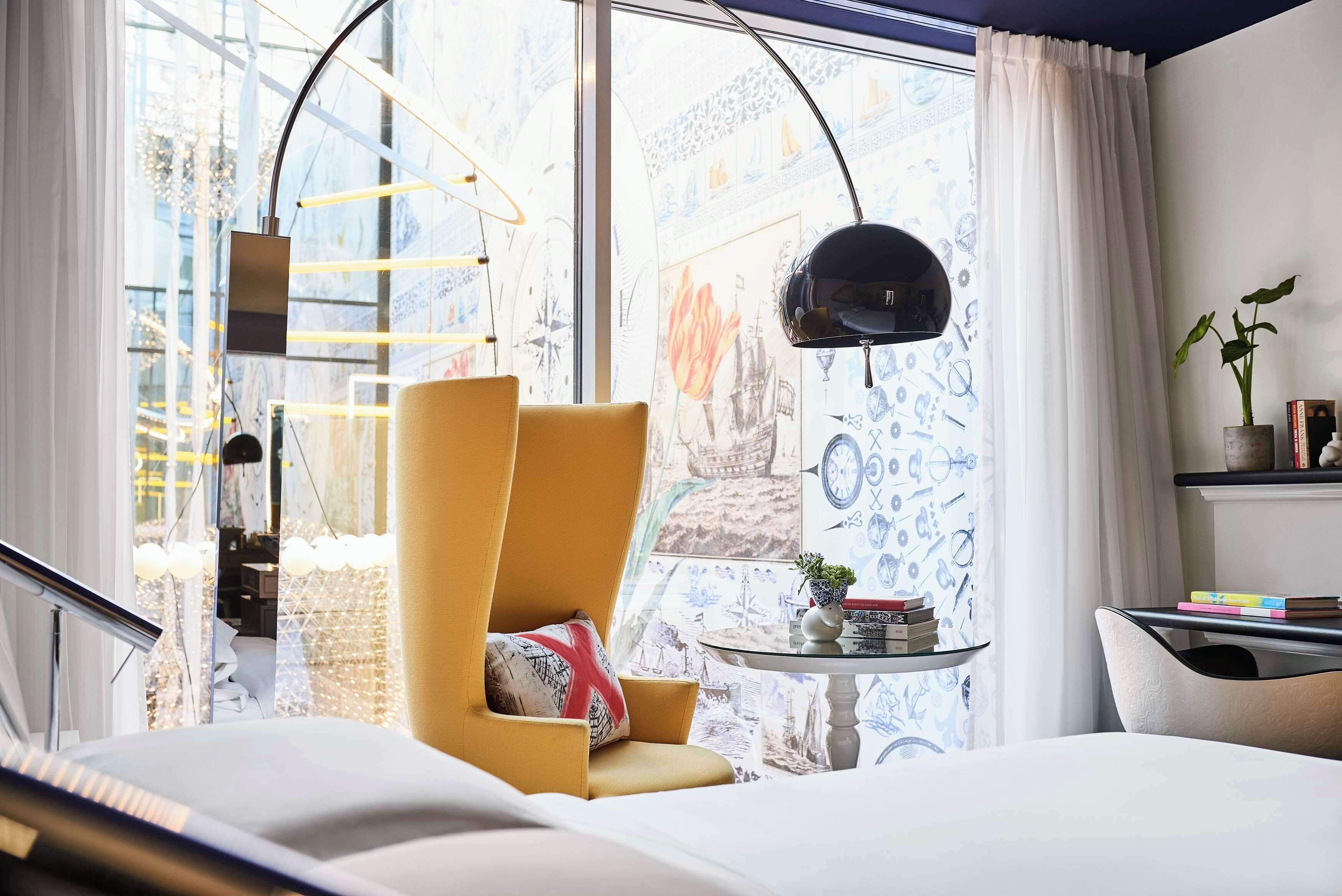 Quarto de hotel moderno com uma janela grande, uma poltrona amarela, uma mesinha redonda e uma luminária pendente preta.