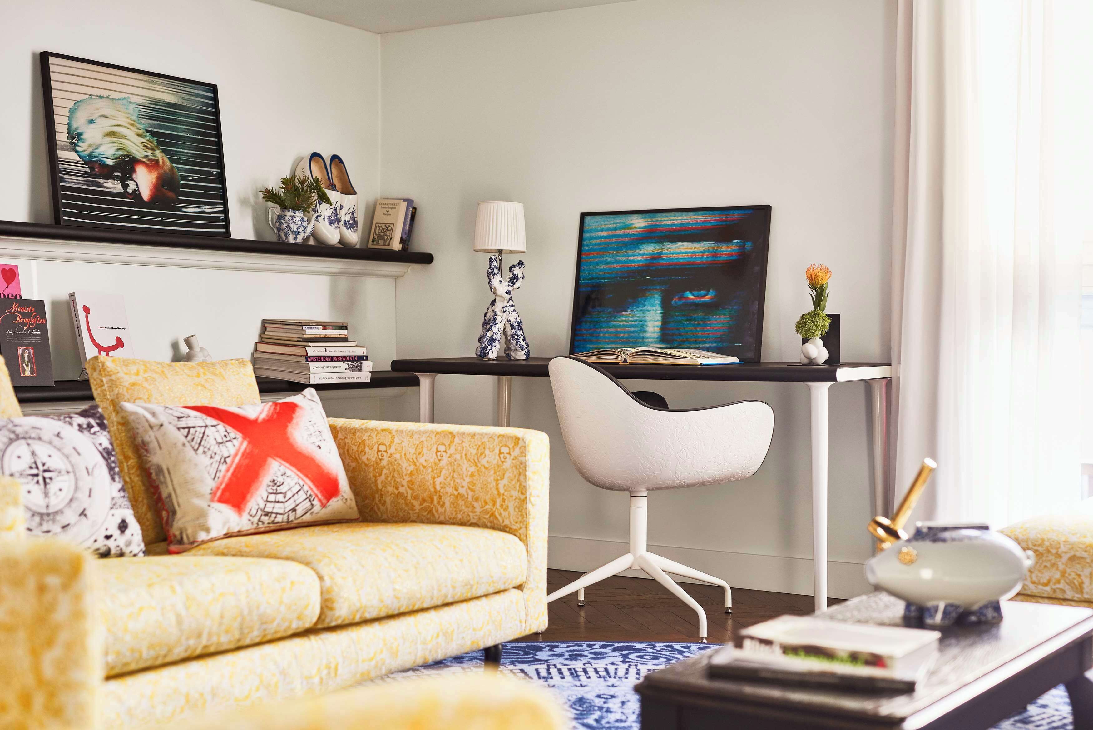 Uma sala de estar moderna com um sofá amarelo, uma cadeira branca, uma escrivaninha com monitor de computador e diversos objetos decorativos.