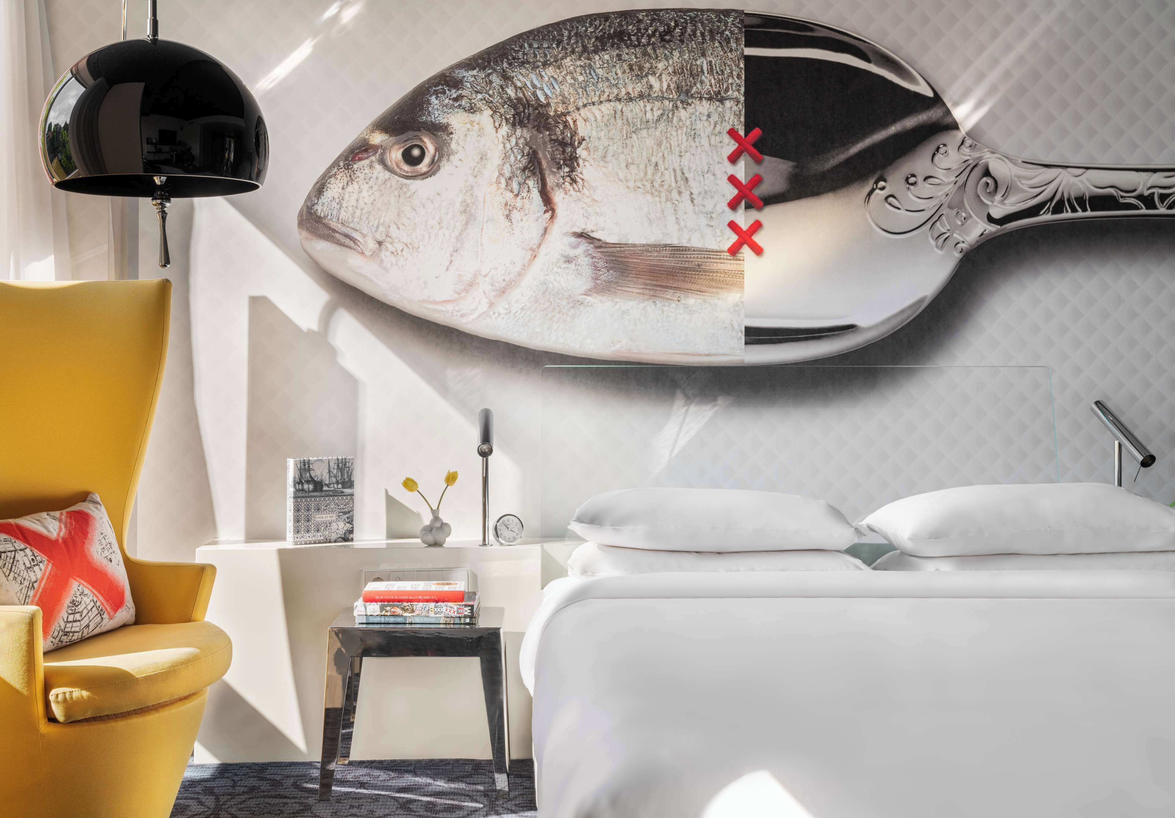 Quarto de hotel moderno com um grande mural de peixes na parede, uma poltrona amarela, uma mesa de cabeceira com livros e uma cama com roupa de cama branca.