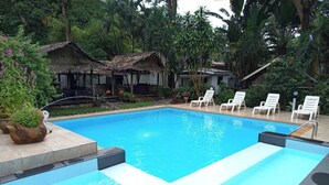 Uma piscina retangular com centro azul translúcido e borda ladrilhada, rodeada por espreguiçadeiras brancas e plantas em vasos.
