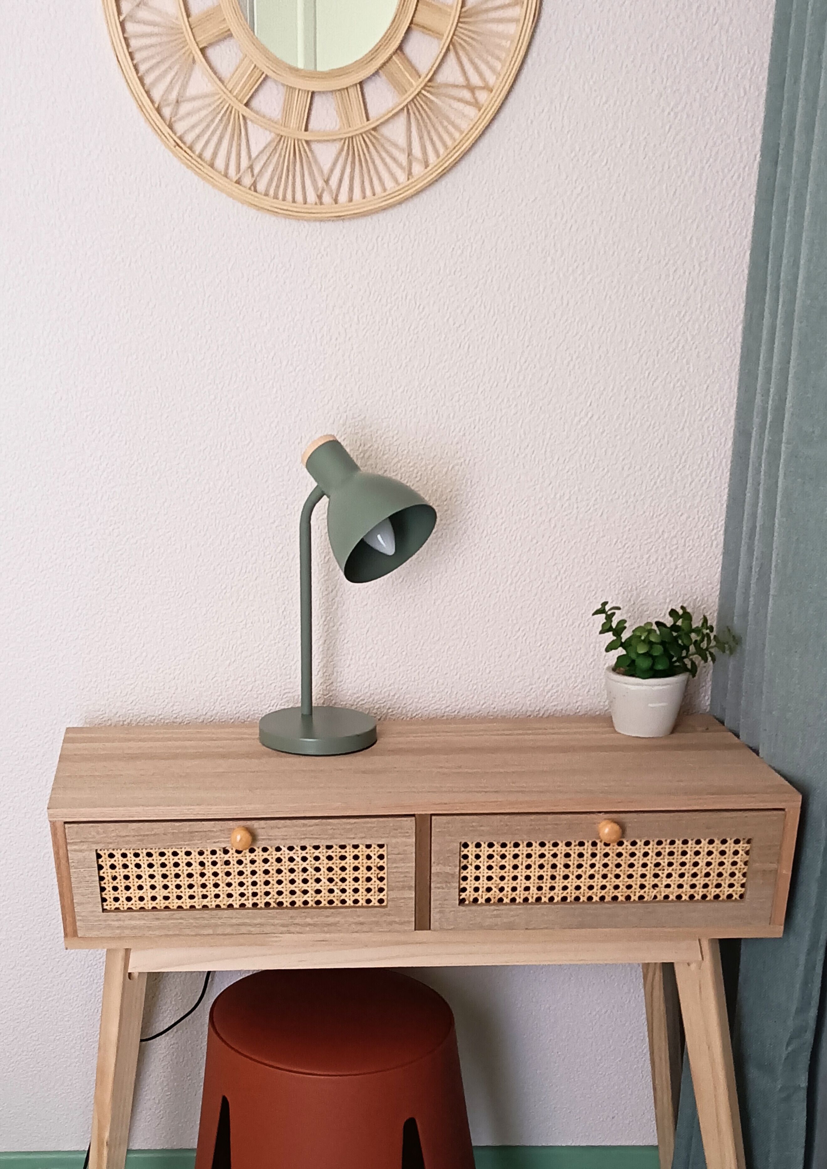 Uma secretária de madeira com uma lâmpada de mesa verde, uma planta em vaso e um espelho decorativo na parede.