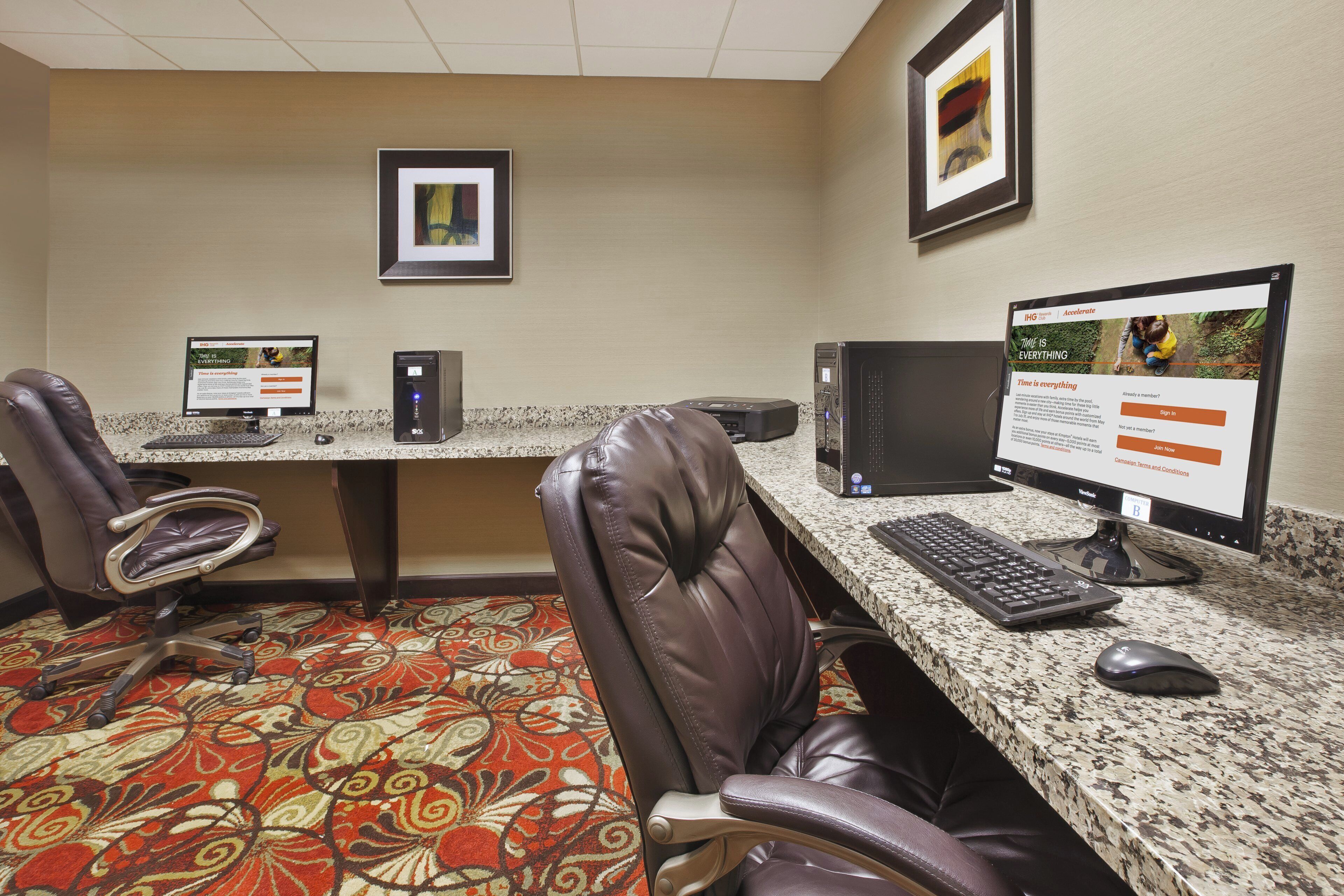 Quarto de hotel com escrivaninha, dois monitores, teclado, mouse e poltrona de couro.