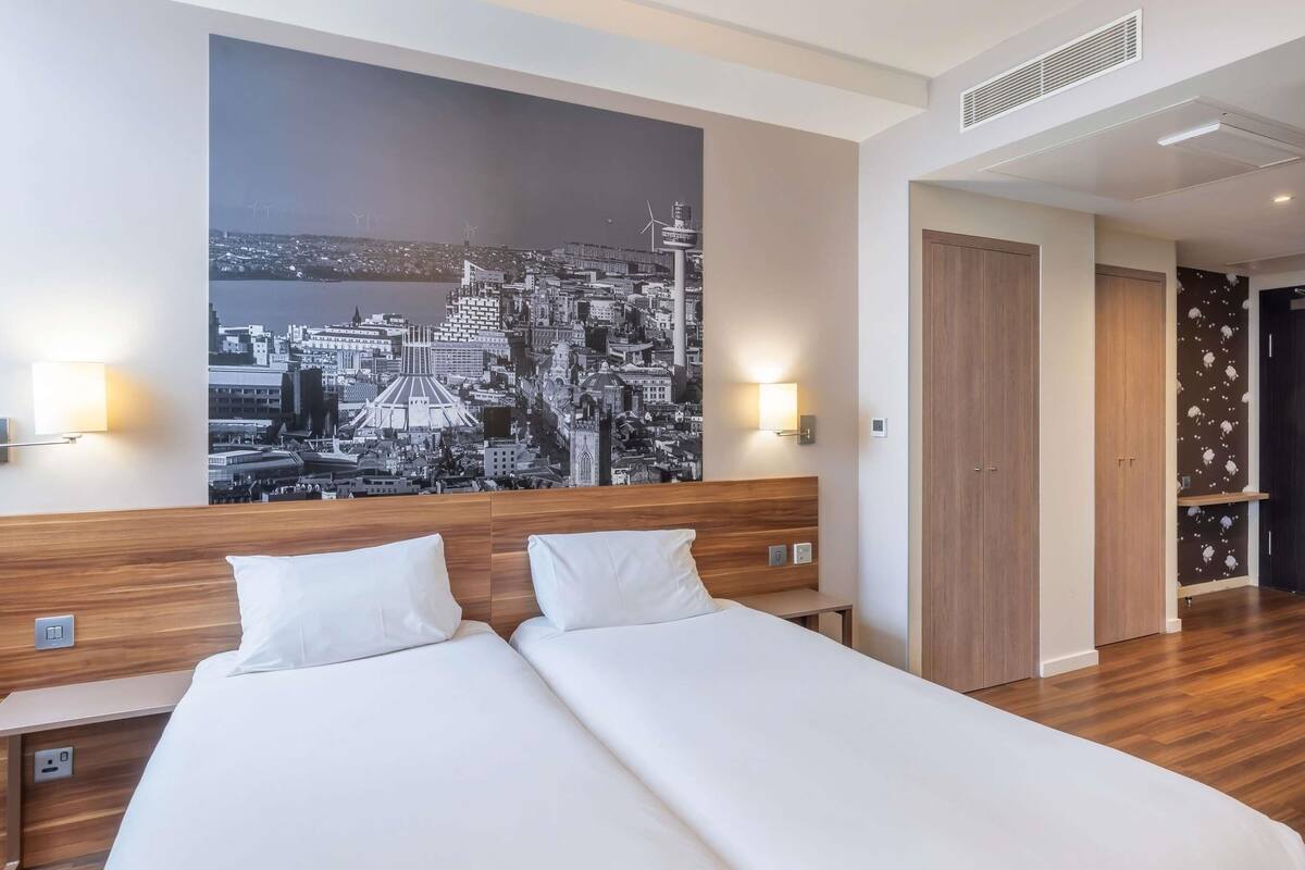 Quarto de hotel com um mural de grande dimensão retratando uma paisagem urbana, cabeceira de madeira, duas camas, abajures e um guarda-roupa.