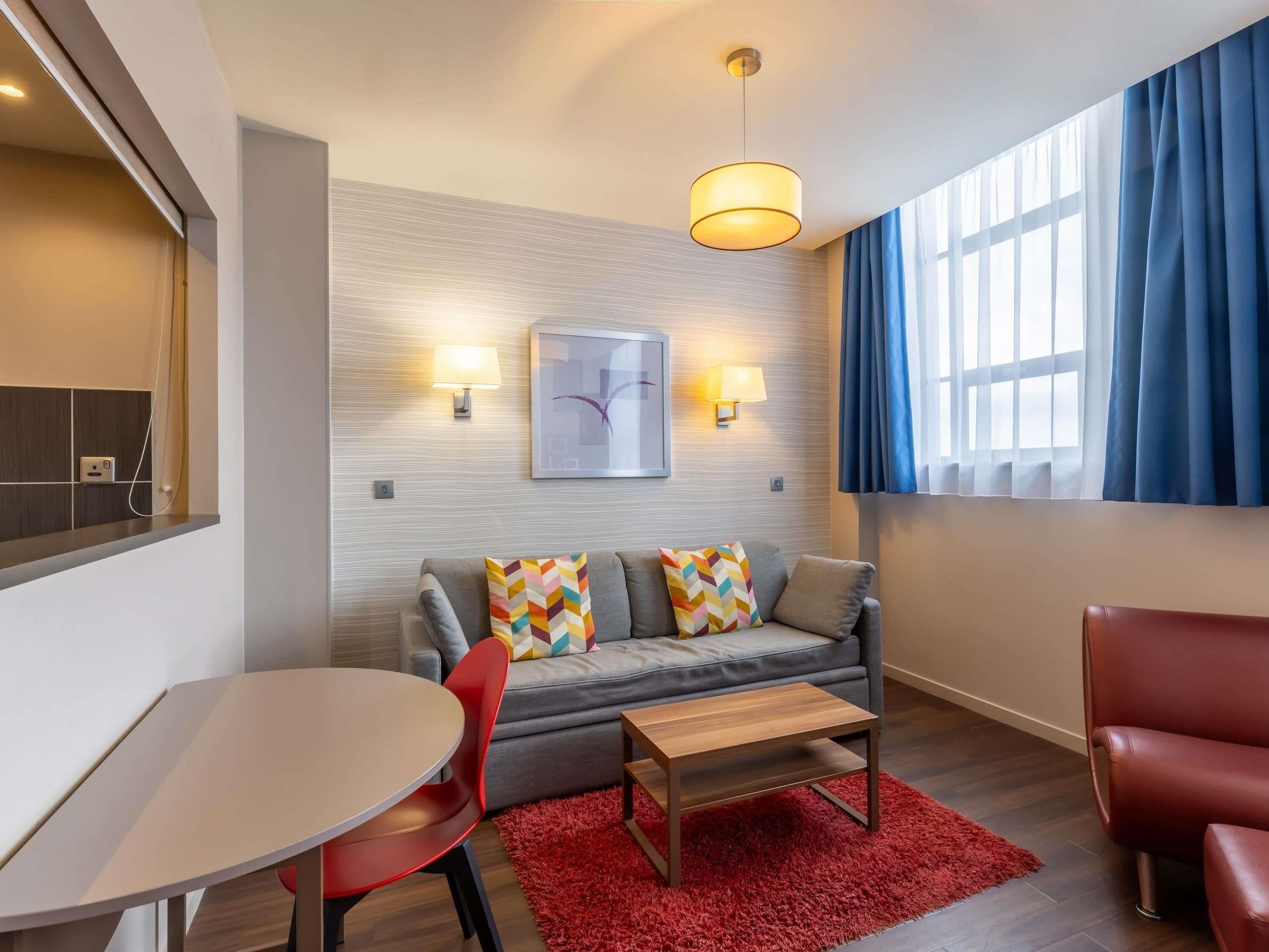 Uma sala de estar moderna com sofá, mesa de centro, mesa de jantar com cadeiras, tapete vermelho e uma janela grande com cortinas azuis.