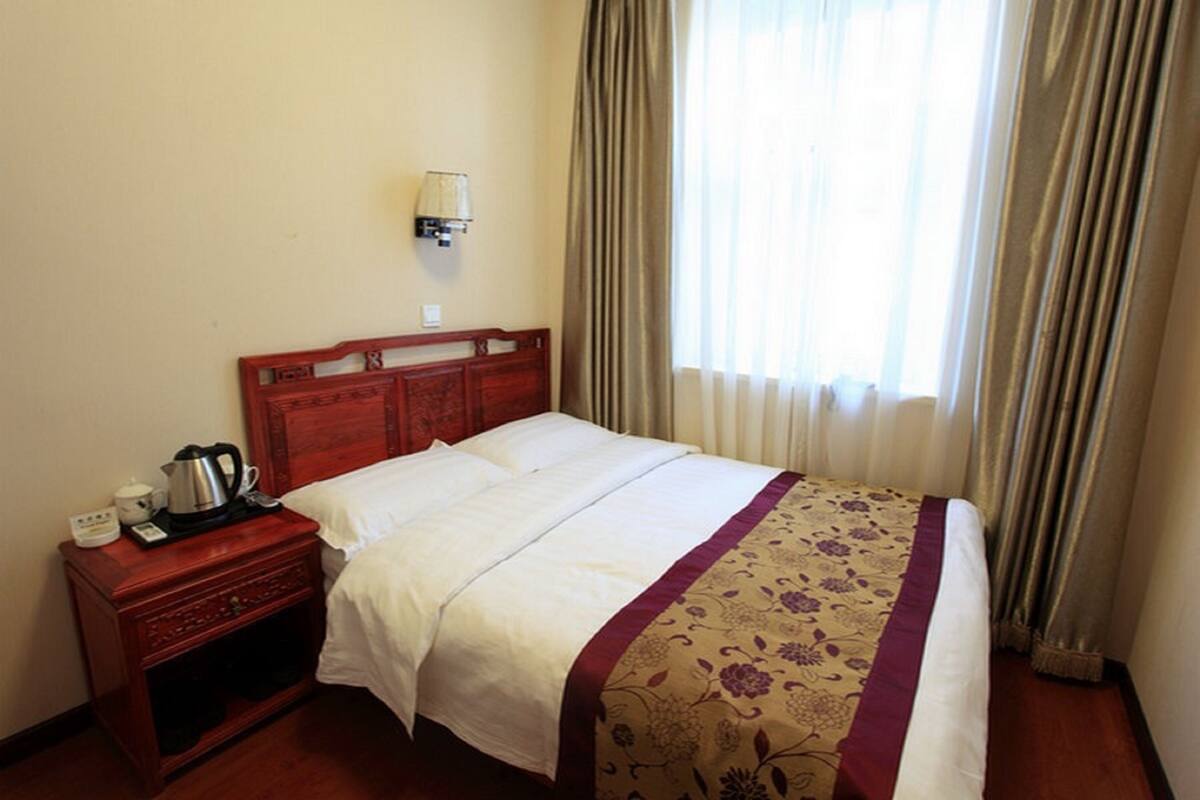 Quarto de hotel com cabeceira de madeira, cama com colcha branca e avelã, mesa de cabeceira com chaleira e xícara, e janela com cortinas.