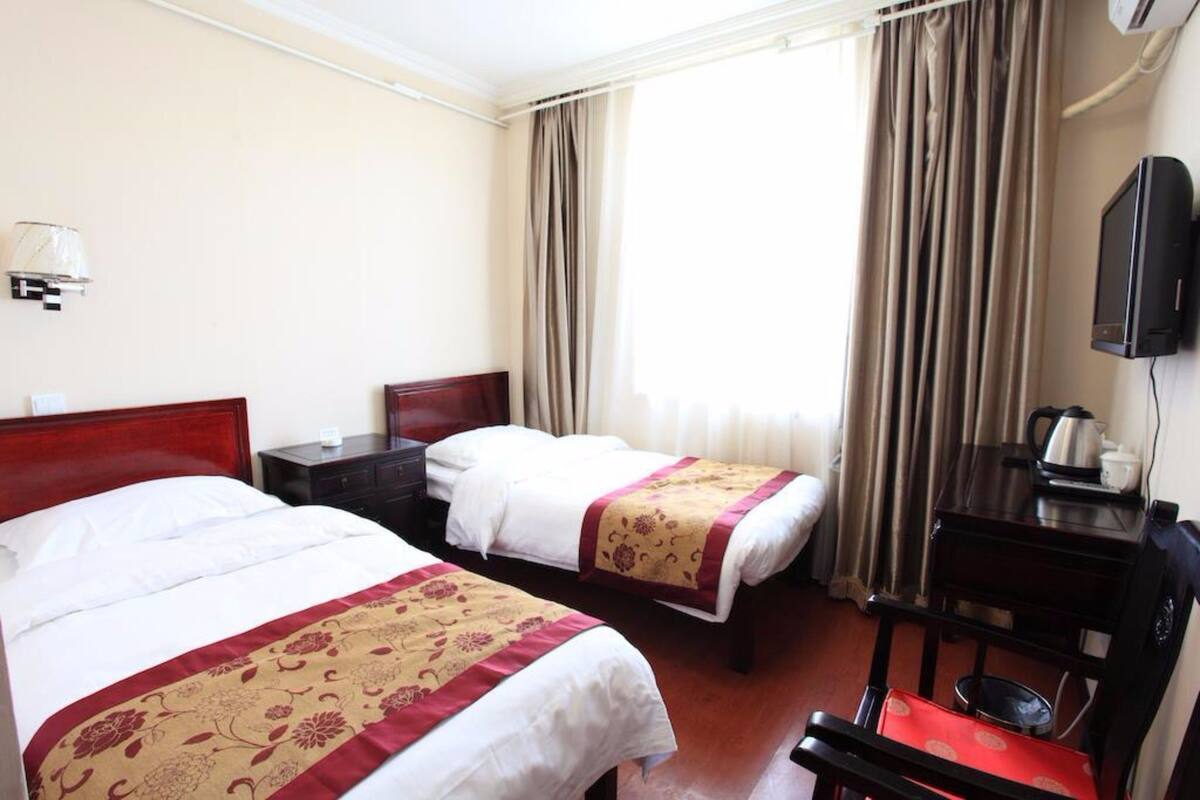 Quarto de hotel com duas camas, uma escrivaninha, uma cadeira, televisão e janela com cortinas.