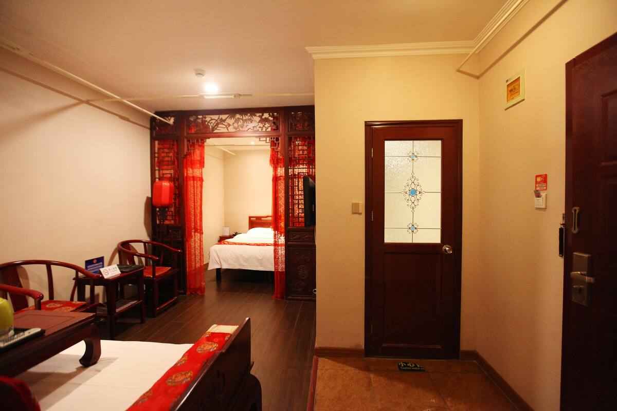 Quarto de hotel com cama de madeira, uma escrivaninha com cadeiras, cortina vermelha e porta com painel de vidro.