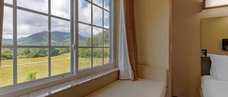 Quarto de hotel com cama, janela com vista para colinas verdes e céu nublado, e luminária fixada na parede.