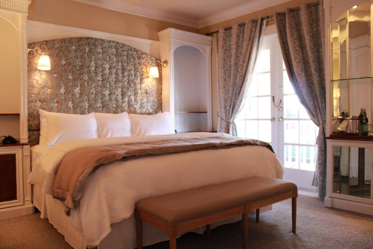 Quarto com uma cama grande, um banco, uma mesinha de cabeceira e um armário de vidro.