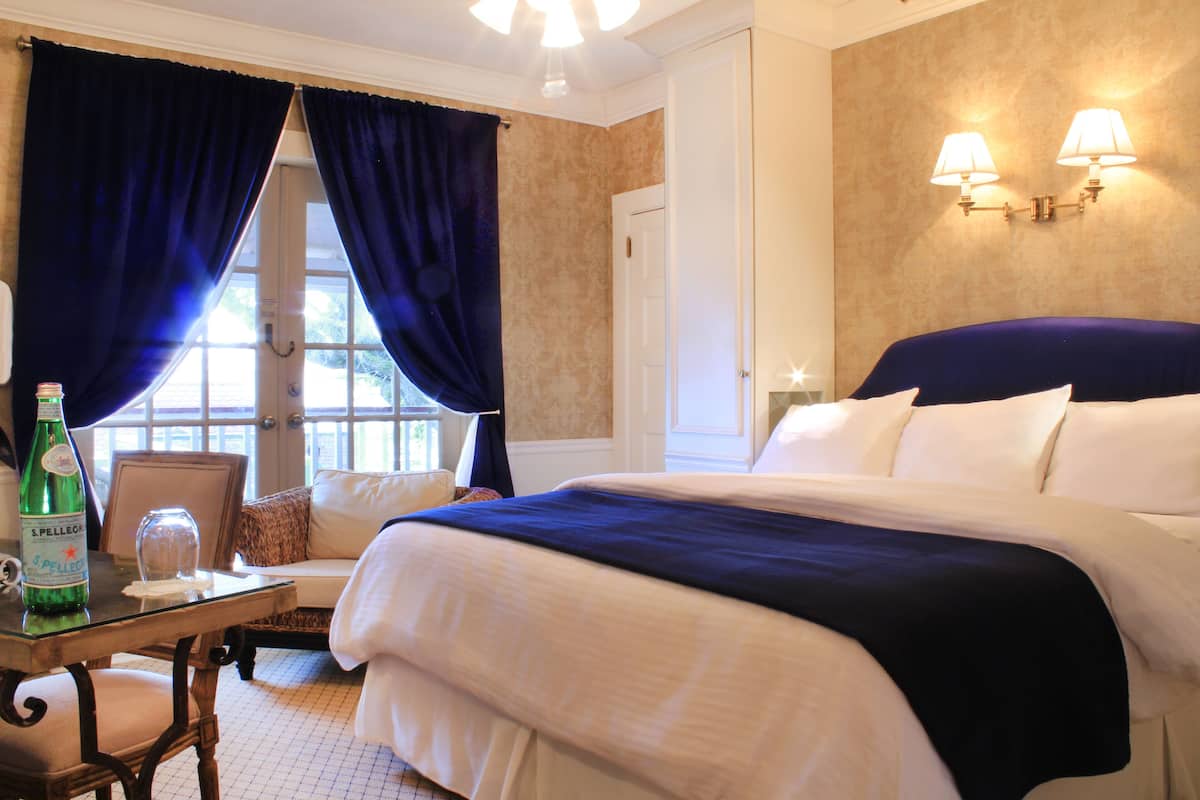 Quarto de hotel com uma cama grande, um espaço de estar com uma cadeira, uma mesa com garrafa e copo, e duas luminárias na parede.