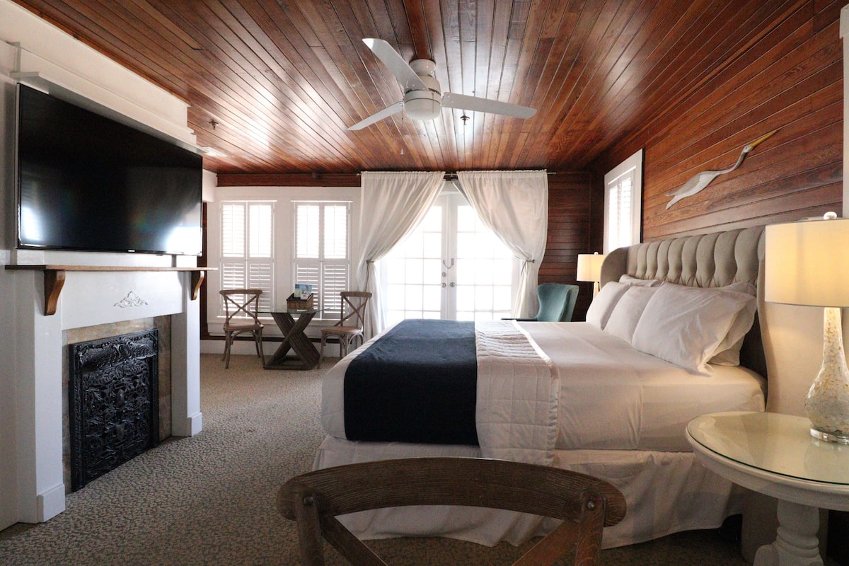 Um quarto com uma cama, televisão, ventilador de teto, mesa com luminária e lareira.
