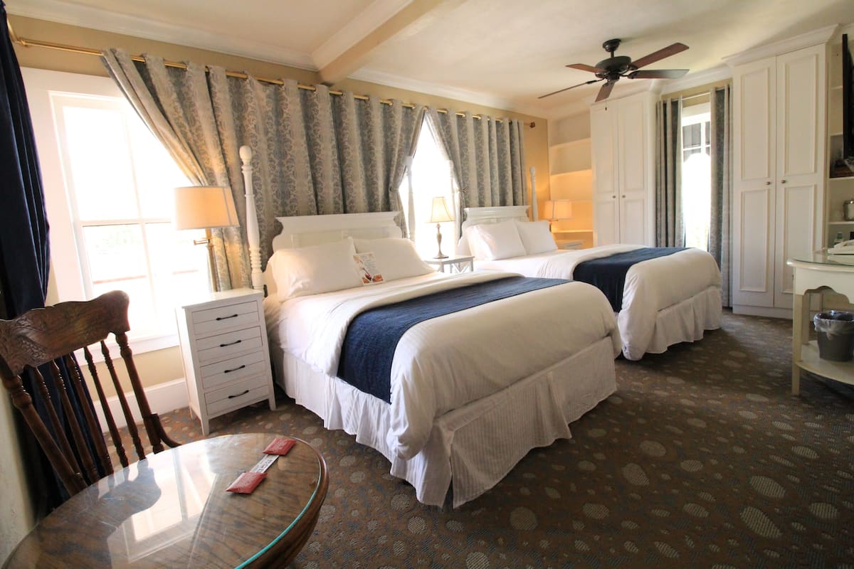 Quarto de hotel com duas camas, uma mesa de jantar, uma cadeira, um ventilador de teto e uma janela grande com cortinas.