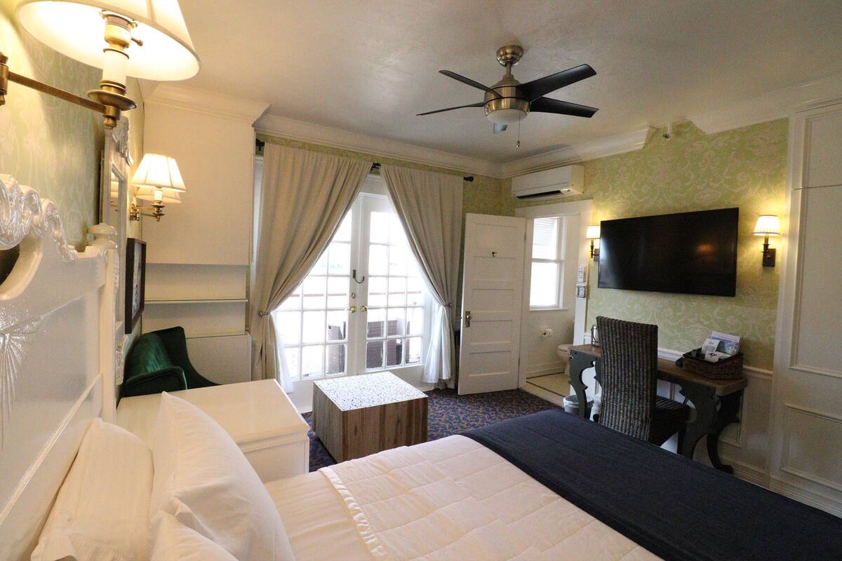 Quarto de hotel com cama, escrivaninha, cadeira, televisão e ventoinha no teto.