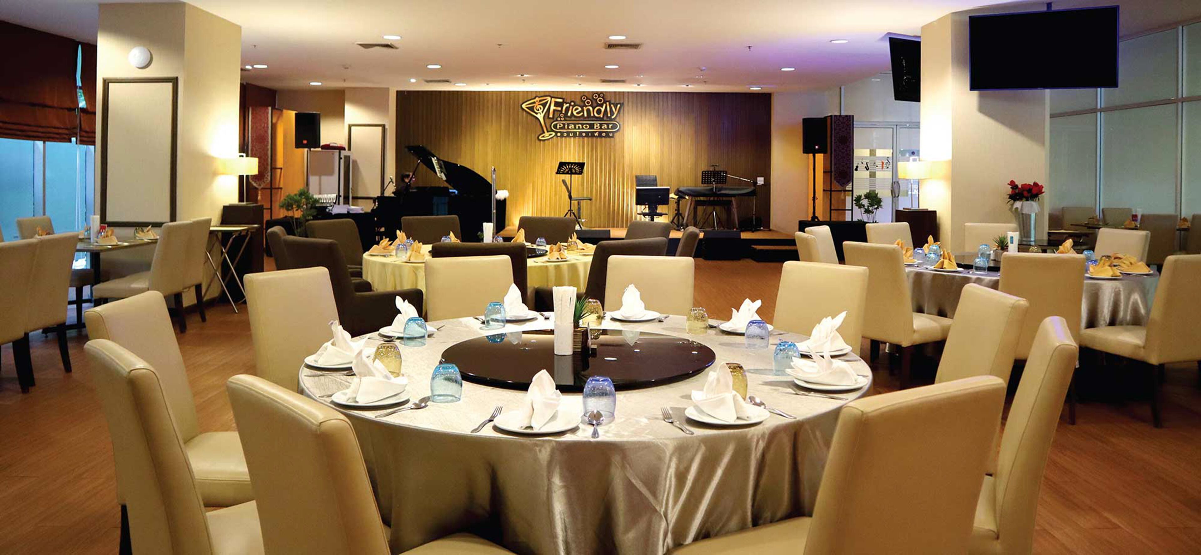 Uma sala de jantar com mesas redondas preparadas para uma refeição, um piano e um palco com instrumentos musicais ao fundo.