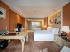 Quarto de hotel com uma cama grande, uma mesa com computador, televisão e banheiro visível por uma porta aberta.