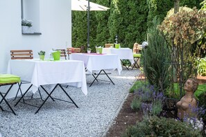 Um pátio externo com mesas e cadeiras, cercado por vegetação e uma estátua.