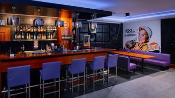 Uma área de bar com um balcão comprido, vários banquinhos e um espaço de estar com cadeiras roxas e uma mesa.