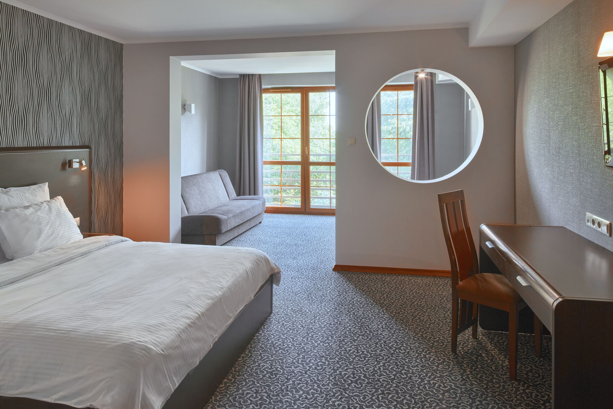 Quarto de hotel com cama, escrivaninha, cadeira, espelho redondo e vista para o exterior pela janela.