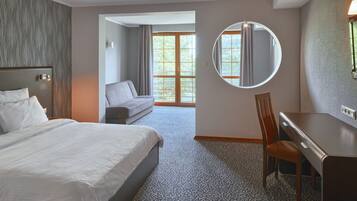 Quarto de hotel com cama, escrivaninha, cadeira, espelho redondo e vista para o exterior pela janela.