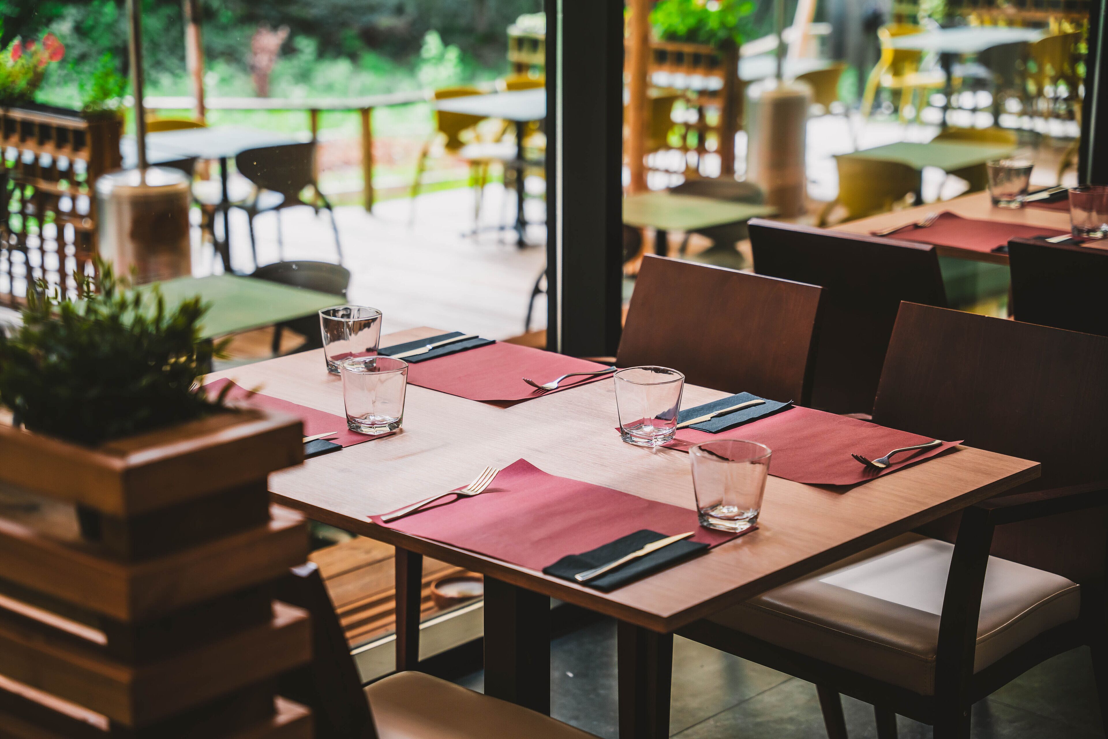 Restaurante com mesas e cadeiras de madeira, jogos americanos vermelhos e copos de vidro transparente.