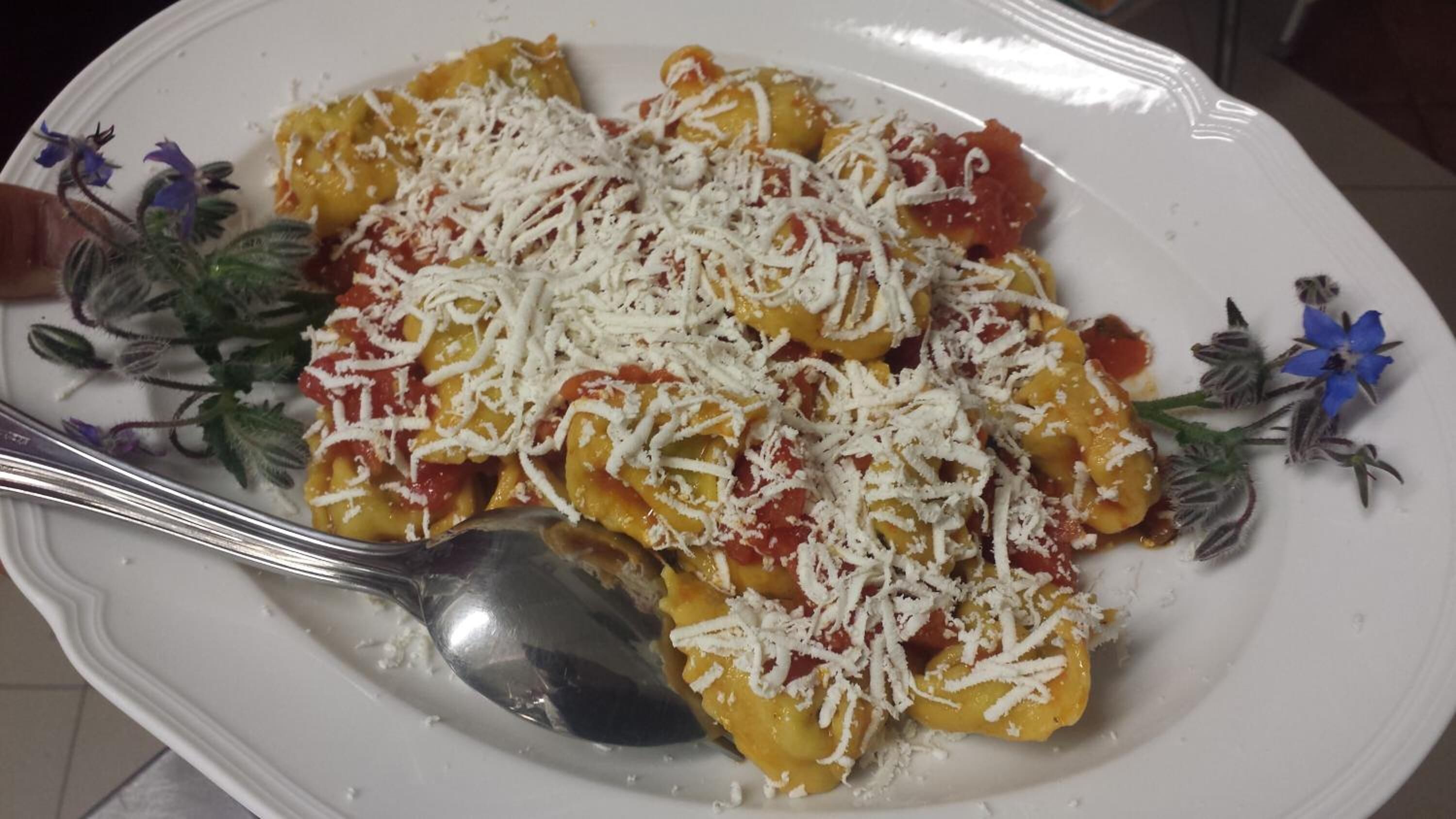 Prato de ravioli com molho de tomate e queijo ralado, acompanhado de uma colher e flores decorativas.