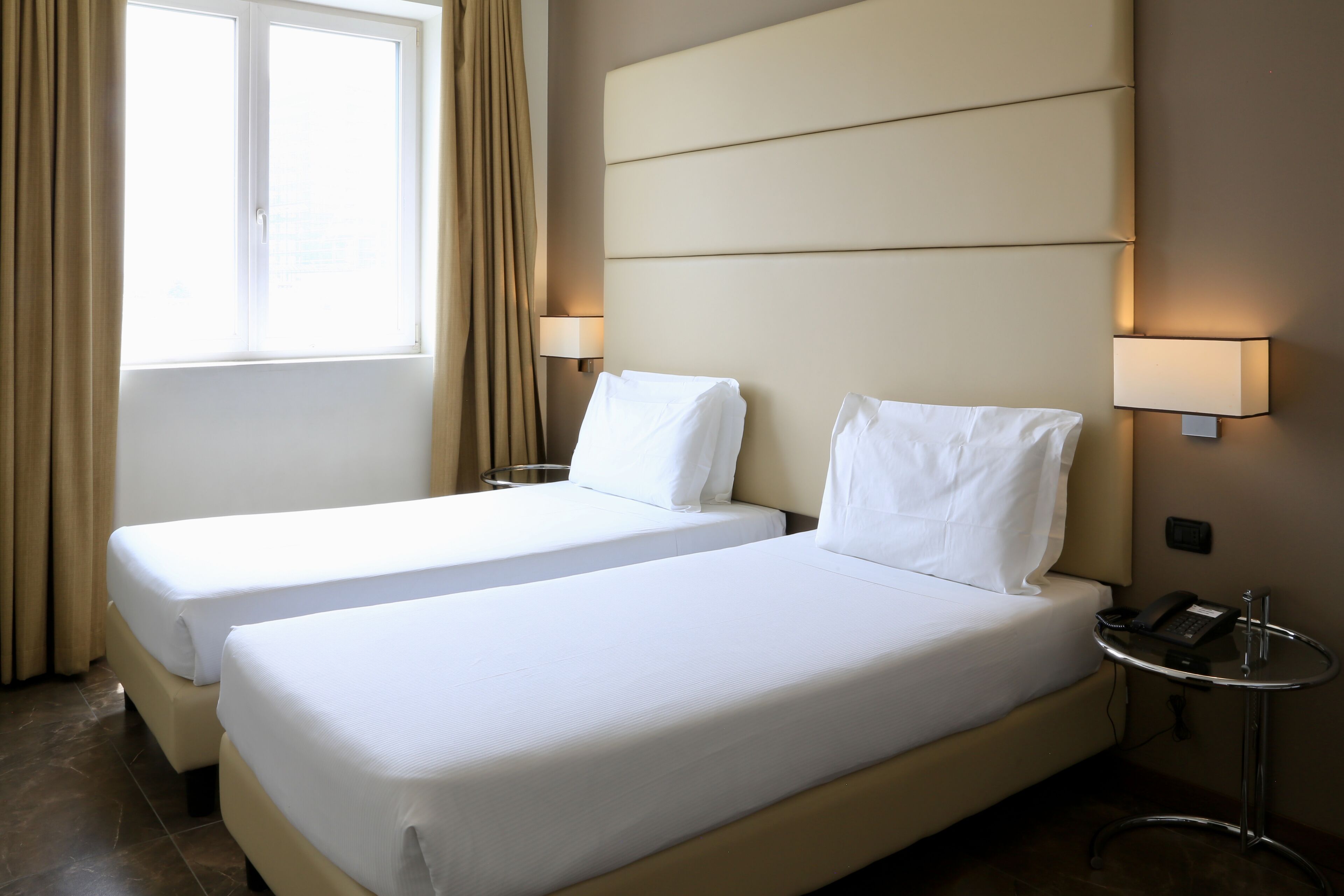 Quarto de hotel com duas camas, janela com cortinas, abajures e uma mesinha com telefone.