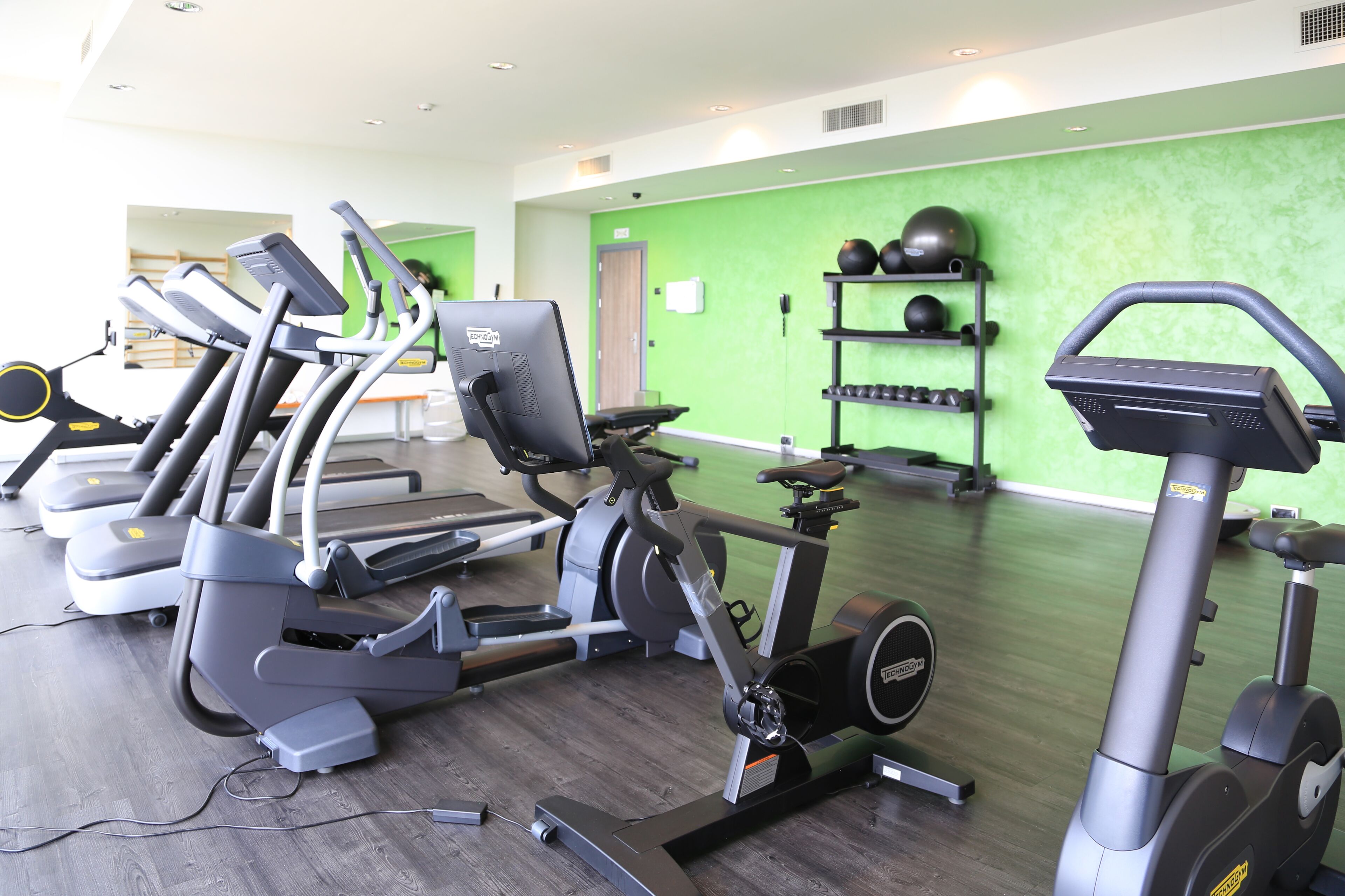 Uma academia com vários equipamentos de exercício, uma parede verde e piso de madeira.