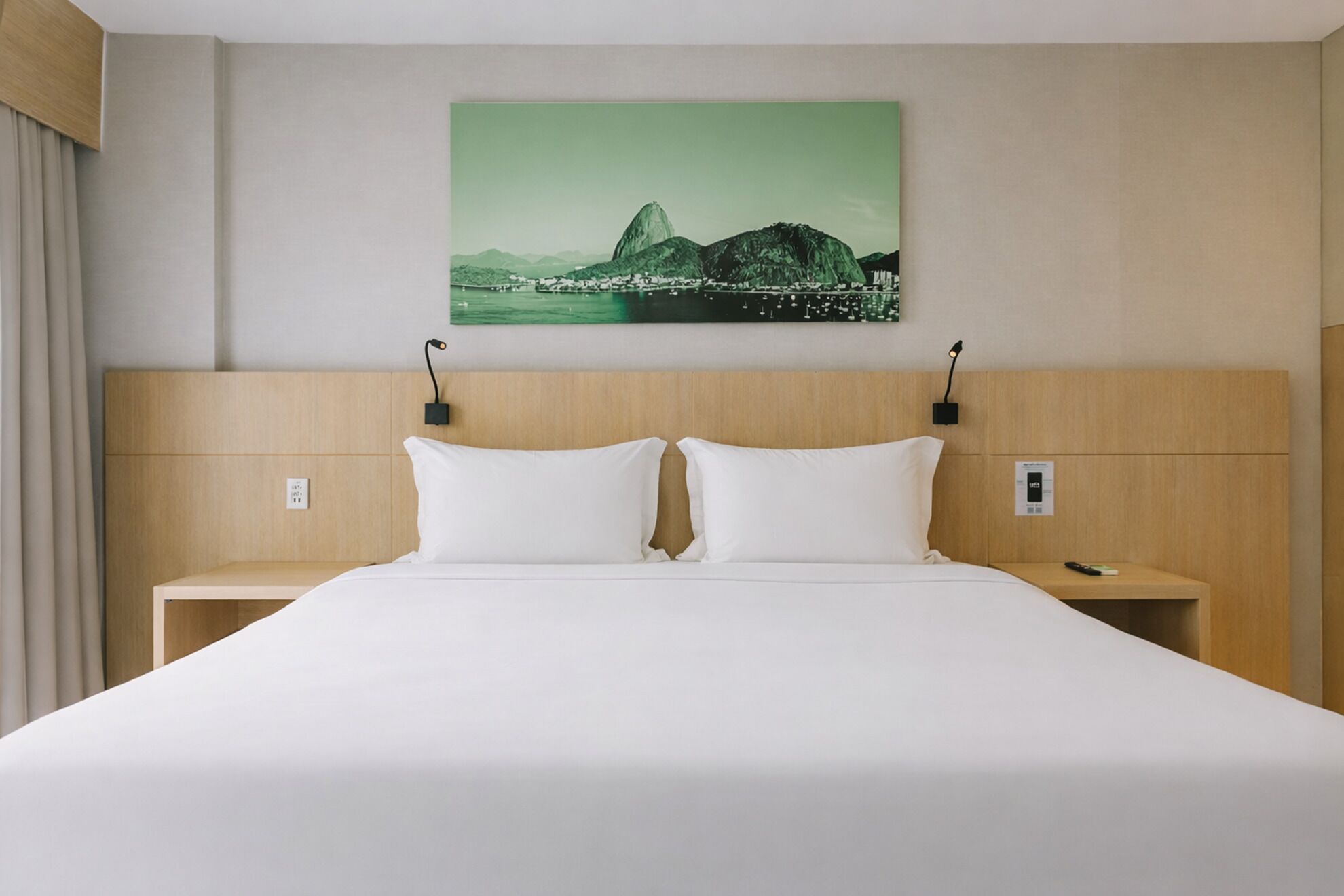 Quarto de hotel com uma cama grande, duas mesas de cabeceira, um criado-mudo, uma luminária na parede e um quadro com paisagem de montanhas.