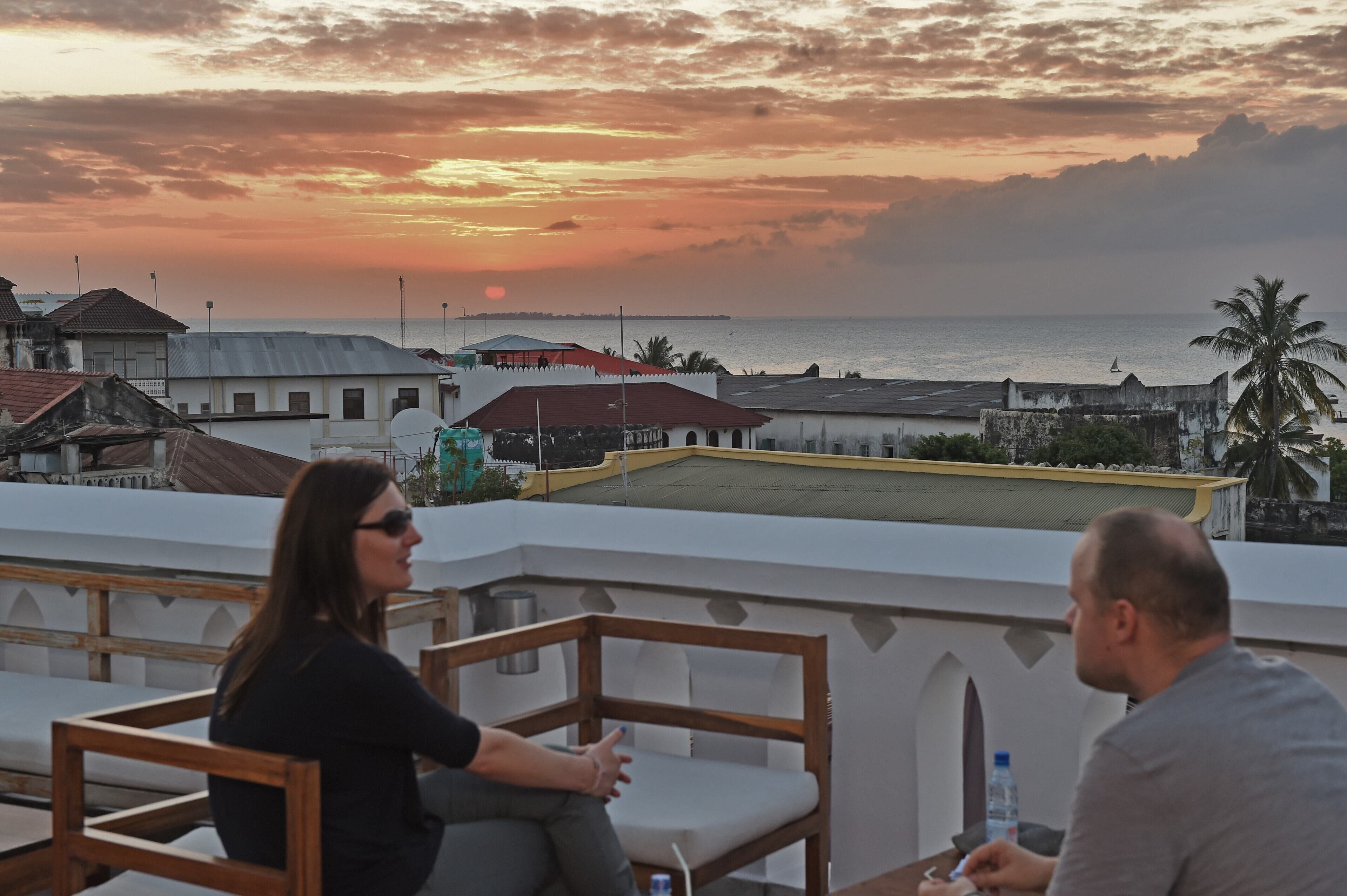 Terraço com mobiliário de jardim e vista para o pôr do sol sobre o mar e edifícios.