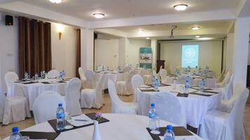 Sala de conferências com várias mesas, cadeiras brancas, garrafas de água e uma tela de projetor exibindo um logotipo.